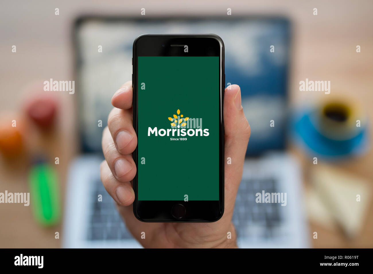 A man looks at his iPhone which displays the Morrisons logo, while sat ...