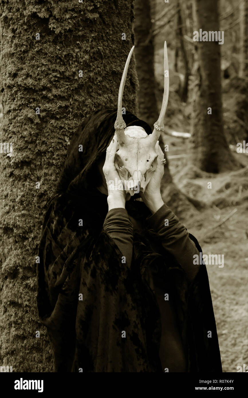 Female witch standing alone in dark woods holding a deer skull - John ...