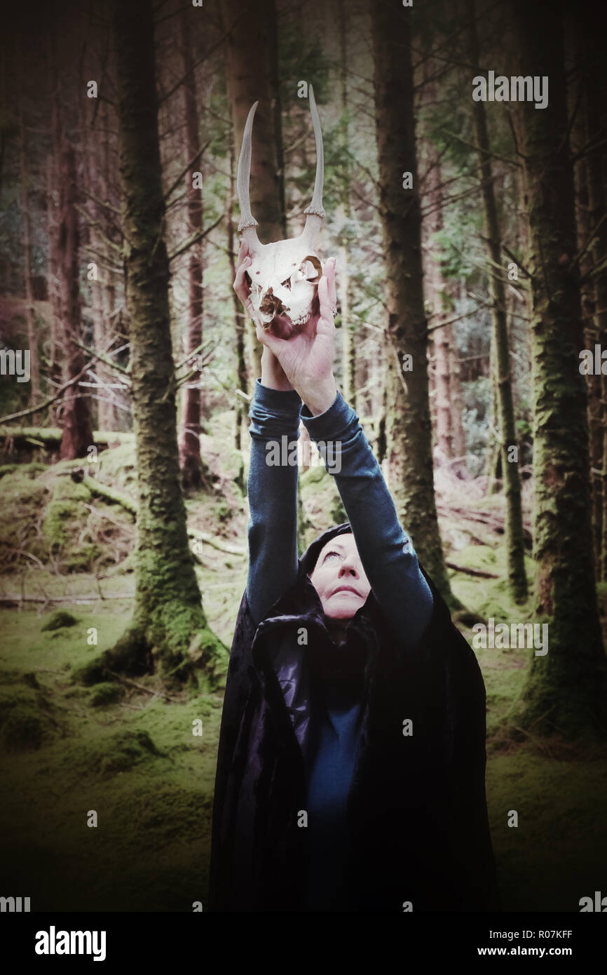 Female witch standing alone in dark woods holding a deer skull - John ...