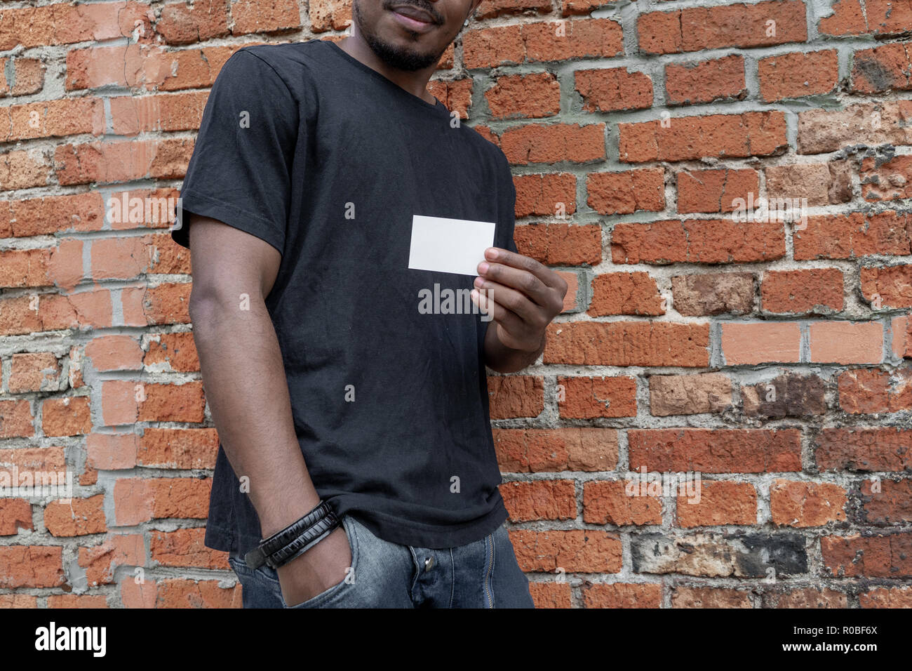 Dark-skinned man in T-shirt showing blank business card on bricked ...