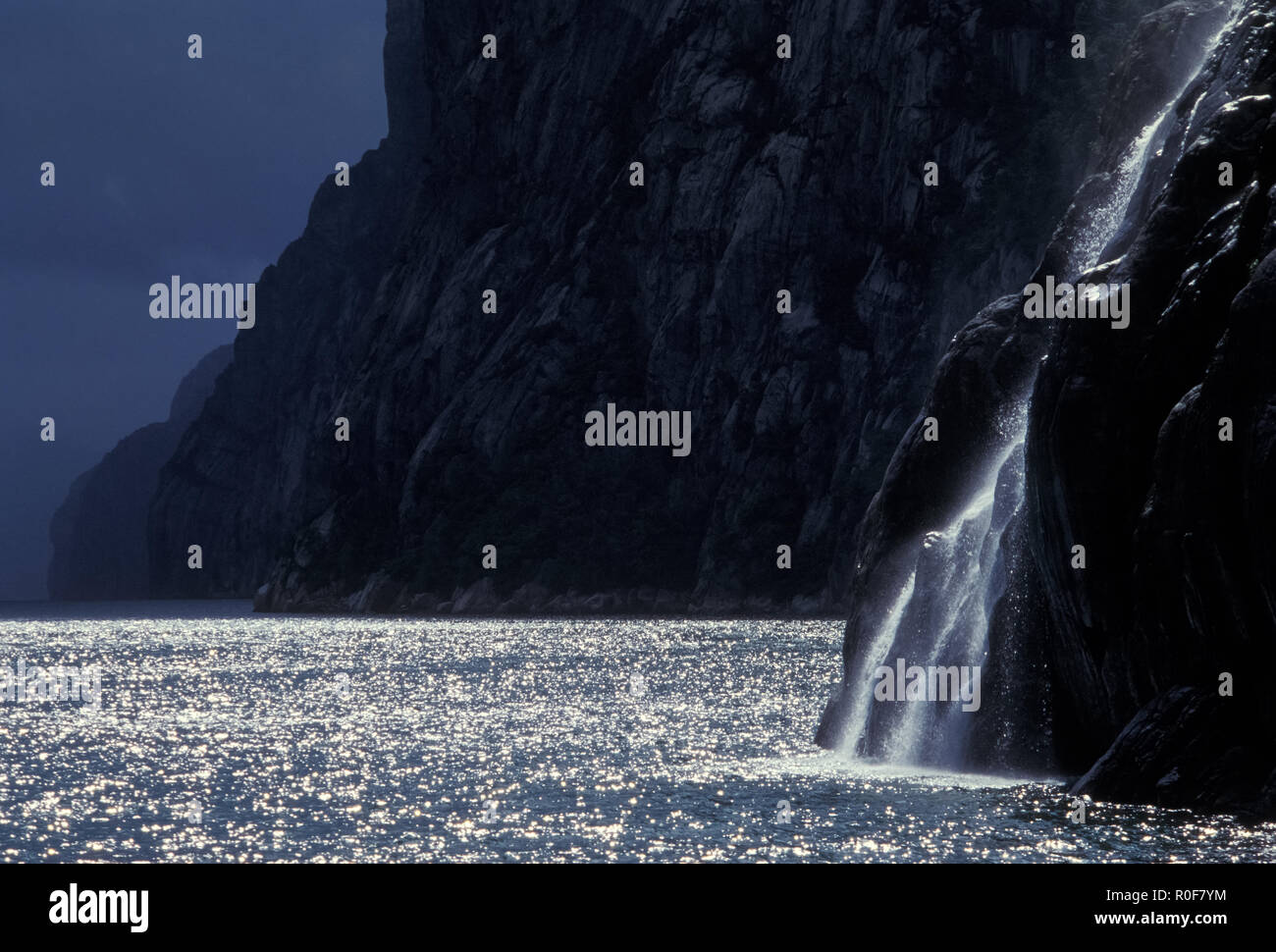 Norwegian fjord with backlit waterfall cascading down the steep cliffs ...