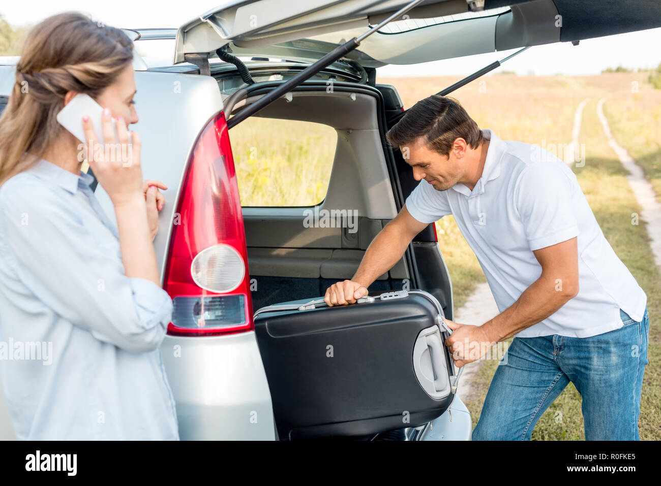 happy adult man packing luggage into car trunk while his girlfriend ...