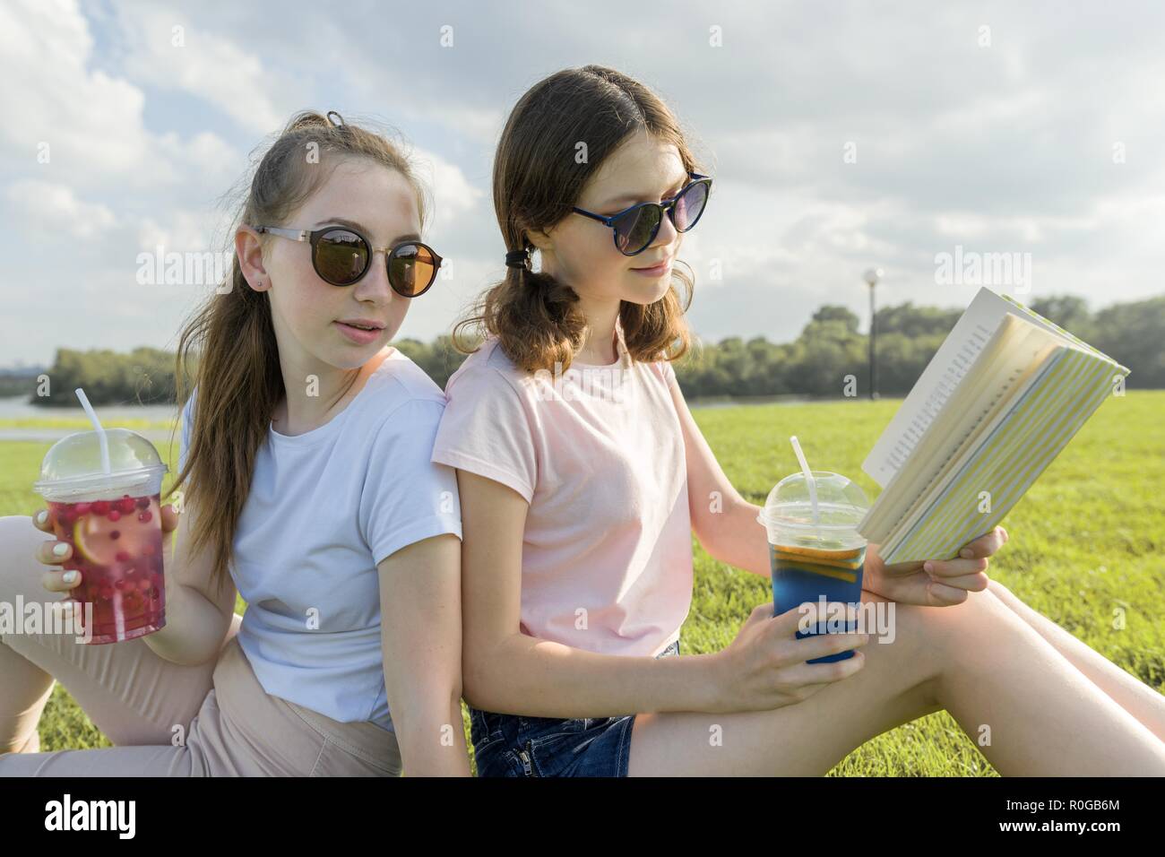 Summer sunny day, two girlfriends teenagers sit on green lawn, drink ...