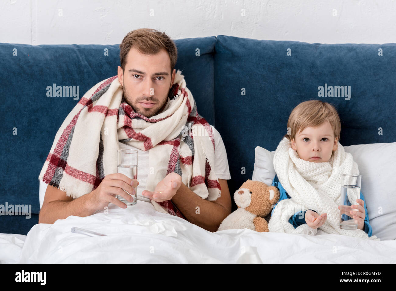 sick father and son with glasses of water and pills sitting in bed and ...