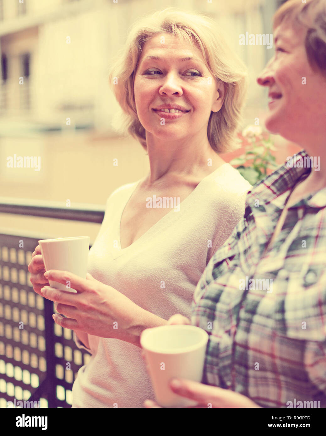 Two smiling elderly women talking on balcony with cup of tea in hands ...