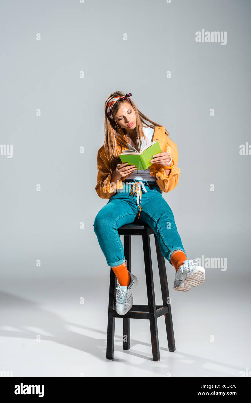 stylish student reading book while sitting on stool on grey Stock Photo ...