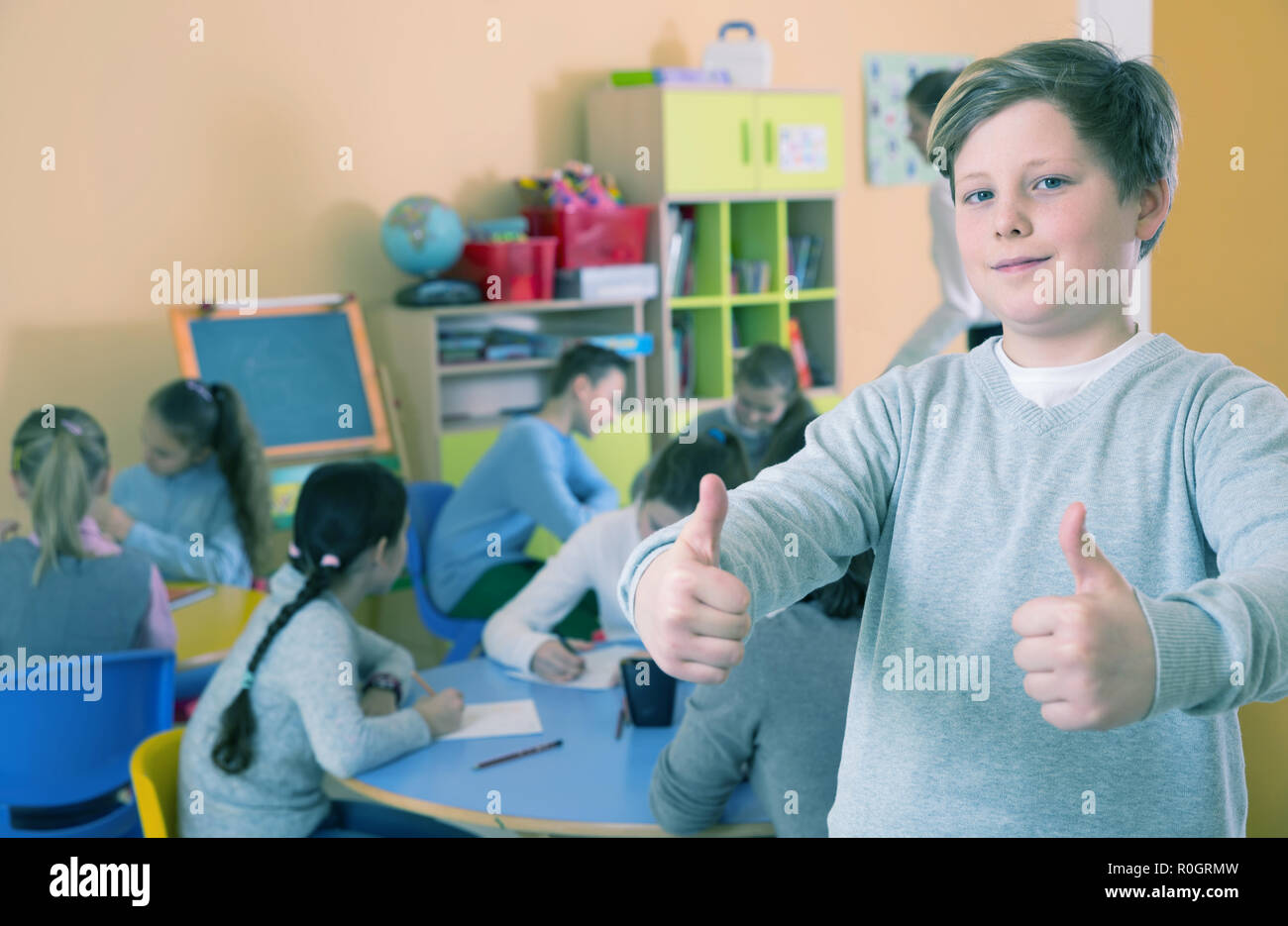 Smiling schoolboy hold thumbs up near children drawing at class Stock ...