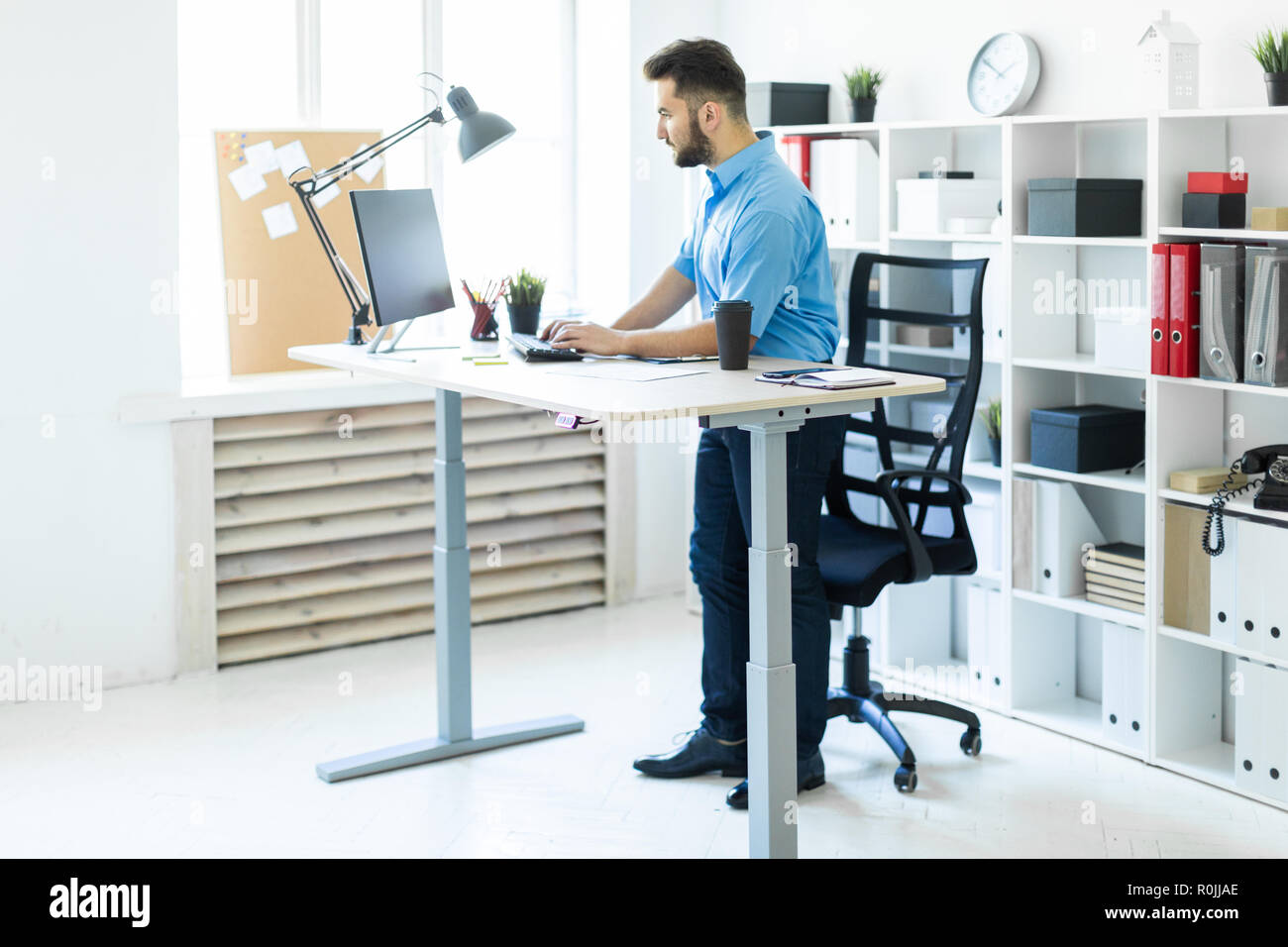 A young man standing in the office and working at the computer table ...
