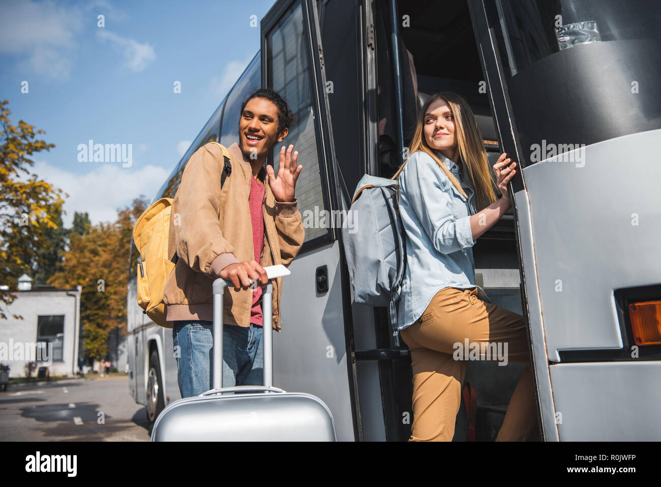 smiling multiracial male tourist with wheeled bag waving by hand while ...