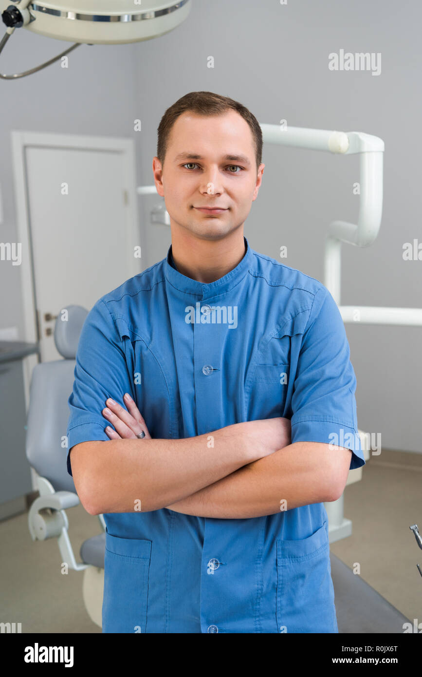 handsome young dentist with crossed arms looking at camera in office ...