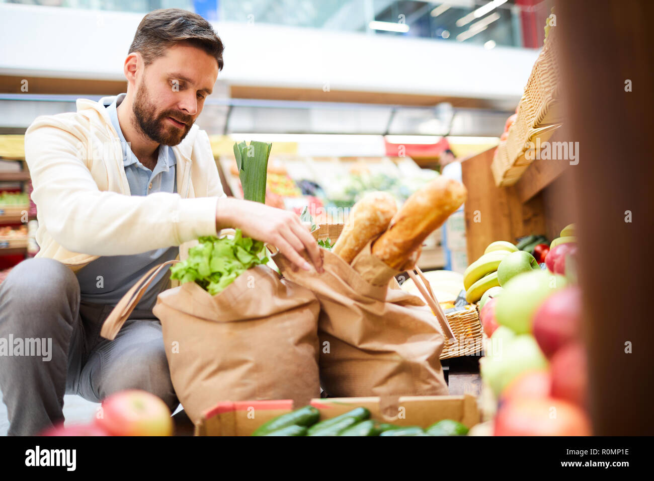 Serious handsome young bearded man in casual clothing packing fresh ...