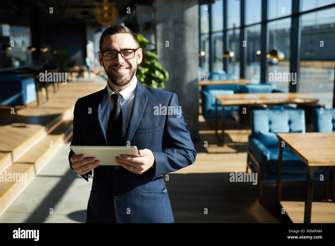 Cheerful man in suit using tablet while working in modern cafe on sunny ...