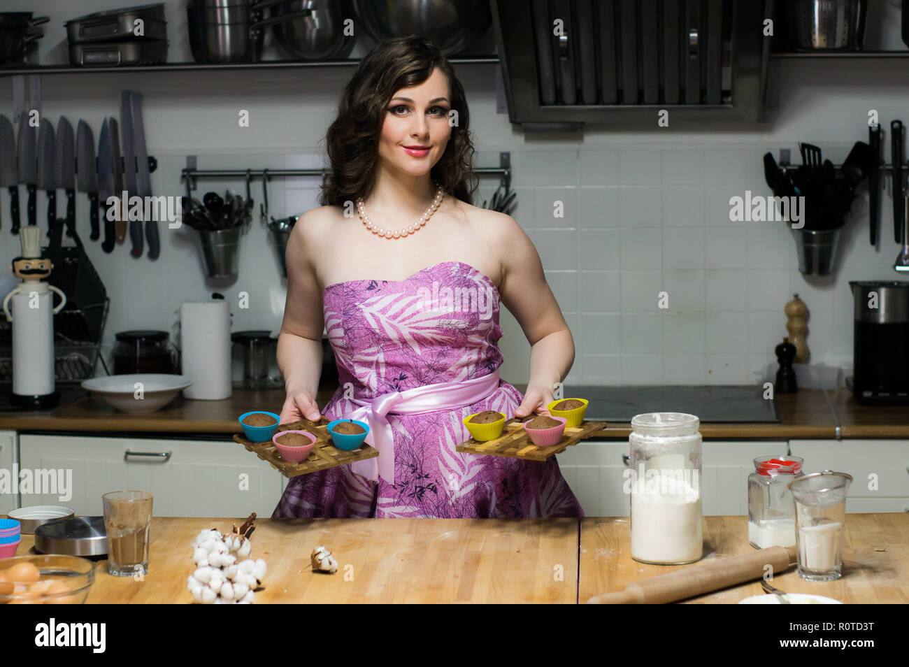 Beautiful girl cooks on kitchen, cupcakes in hands, looks to the camera ...