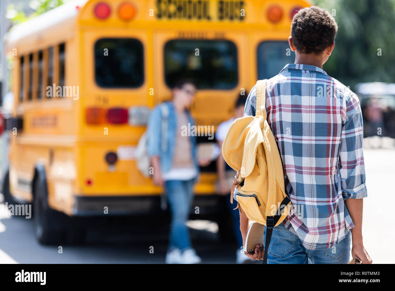 rear view of teen schoolboy walking to classmates leaning on school bus ...