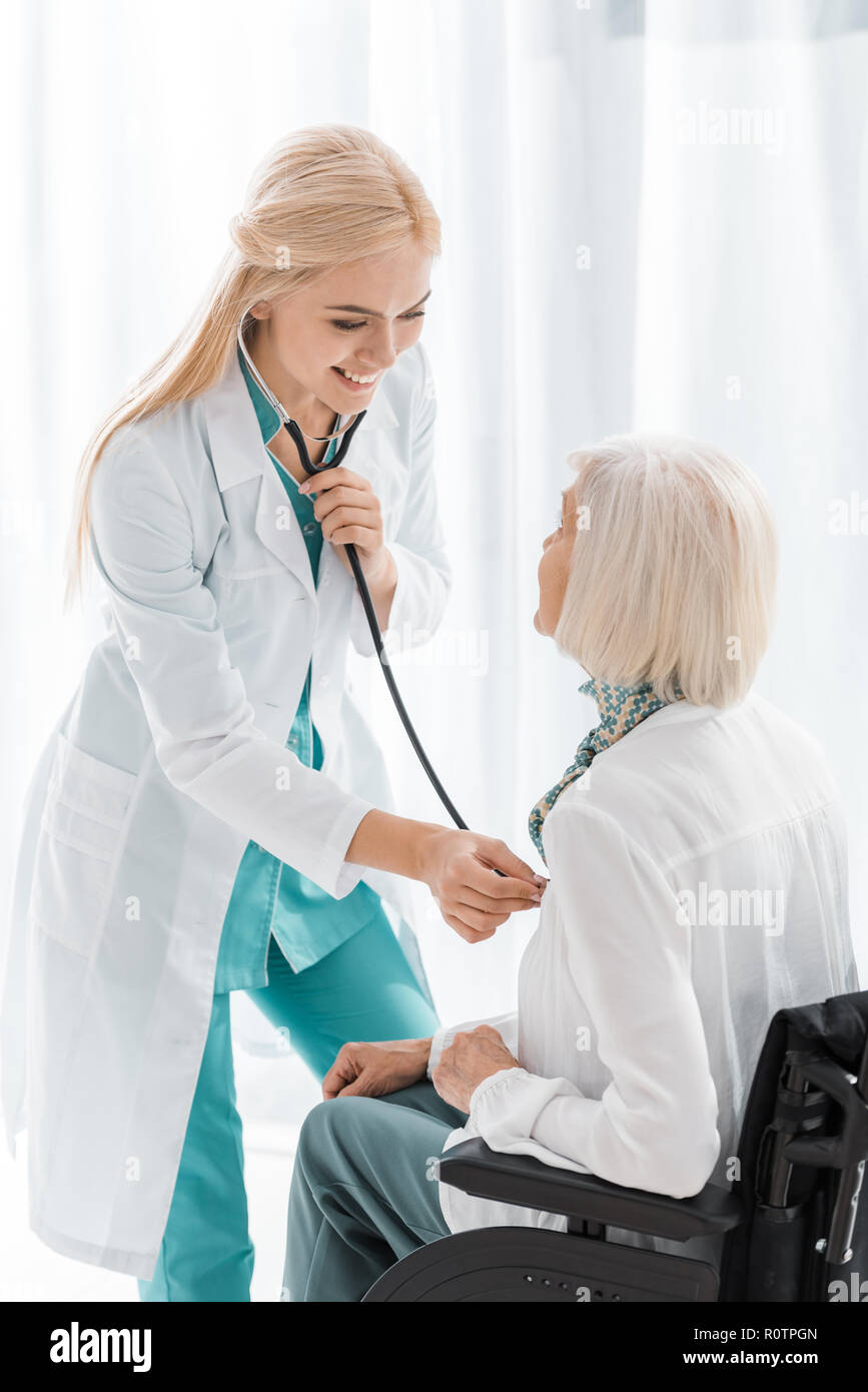 smiling female doctor examining with stethoscope disabled senior woman ...