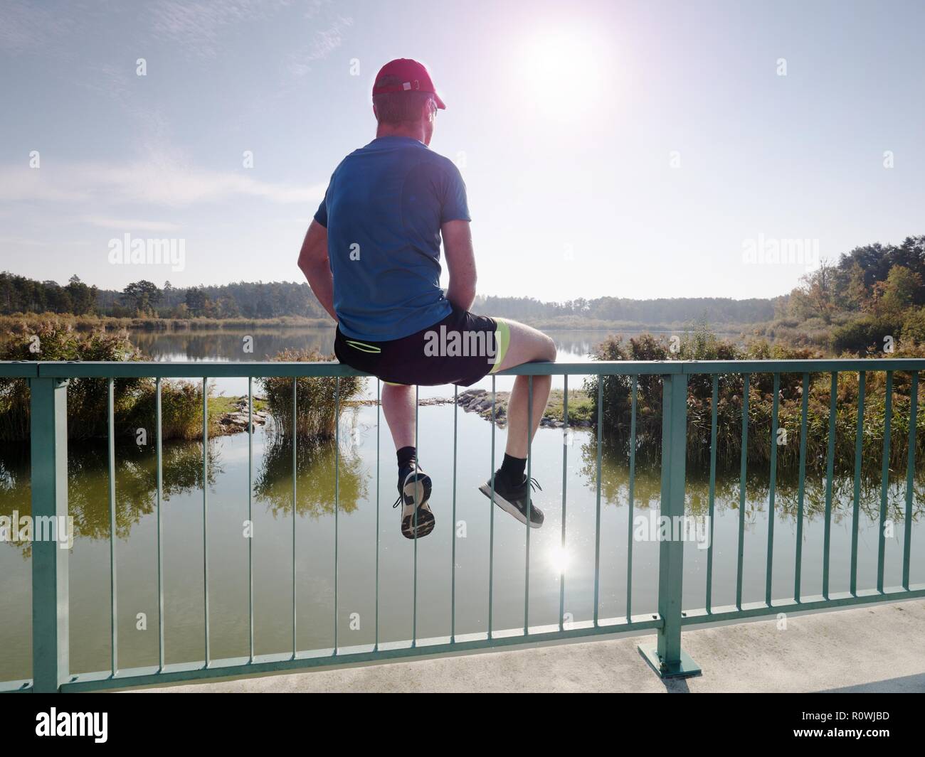 Single Man in sport clothing sit on bridge handrail and watch over lake ...