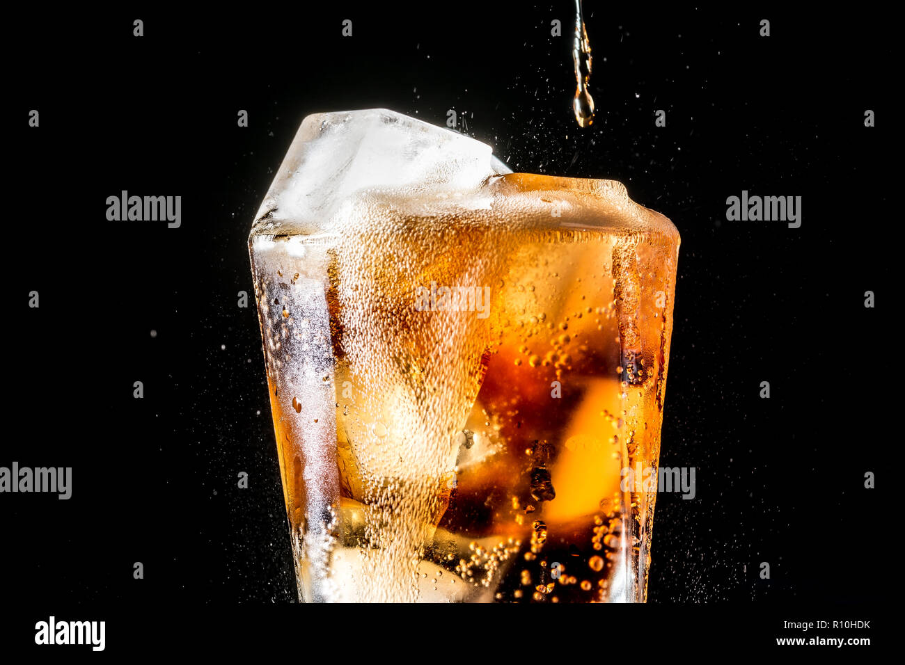 Stream of soda being pouring into a glass with soda and ice cube on black background, closeup ...