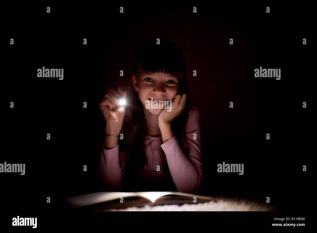 little girl is reading a book with a flashlight in a dark room at night ...