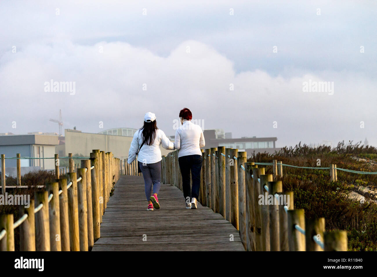 Two middle-aged women walk on a boardwalk on a cloudy day. Back view ...