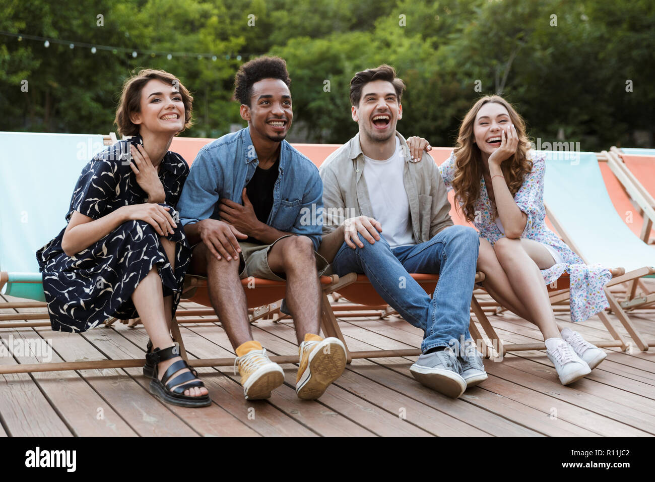 Image of laughing young group of friends sitting outdoors looking aside ...
