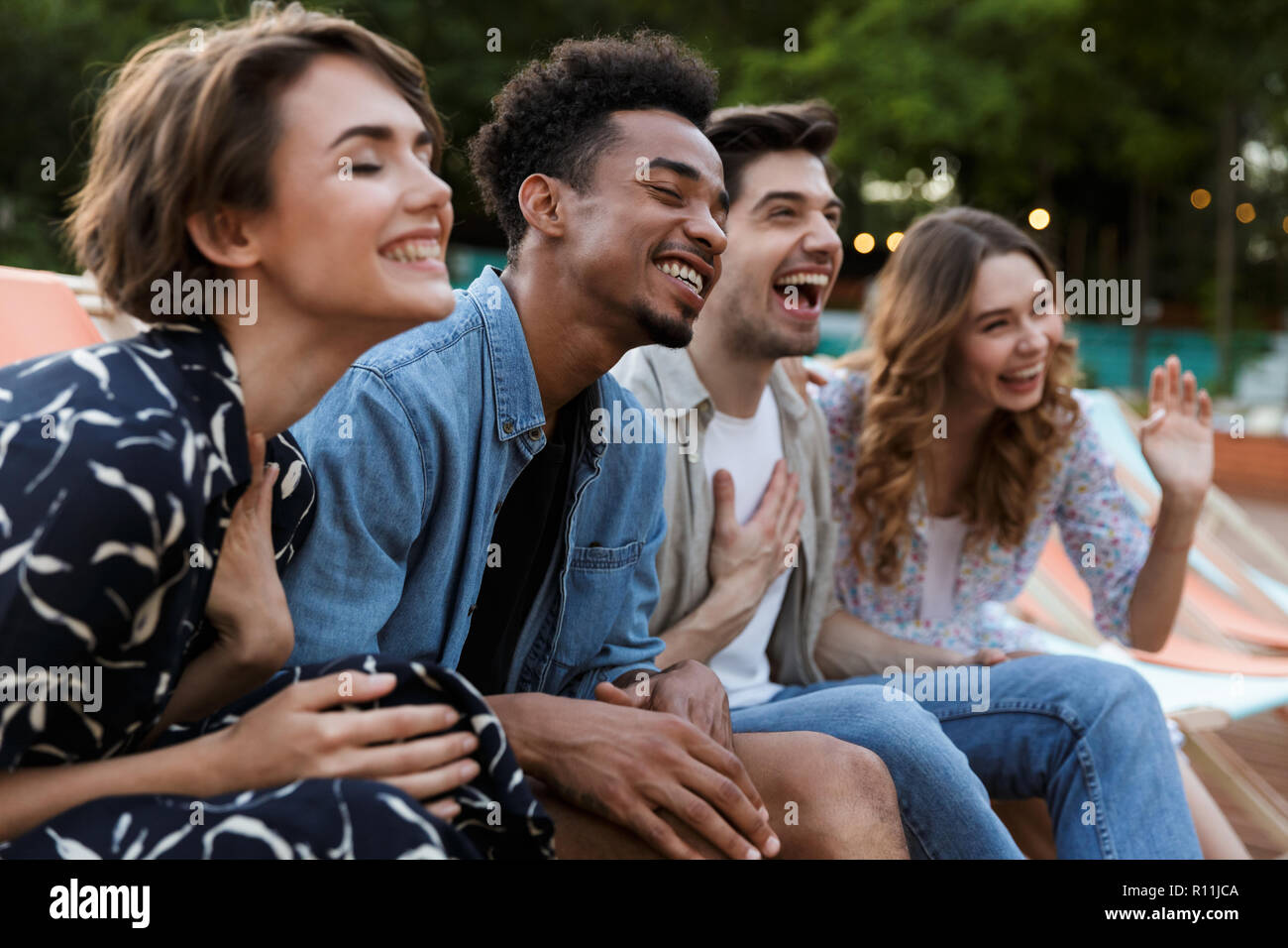 Image of laughing young group of friends sitting outdoors looking aside ...
