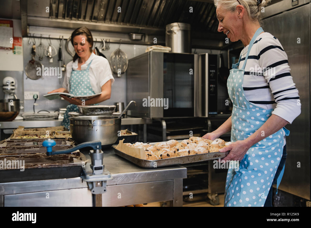 Mature female baker carrying a tray of fruit scones towards the worktop ...