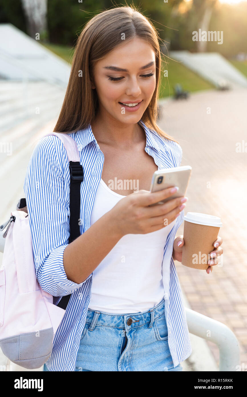 Smiling young girl with backpack holding takeaway coffee while using ...