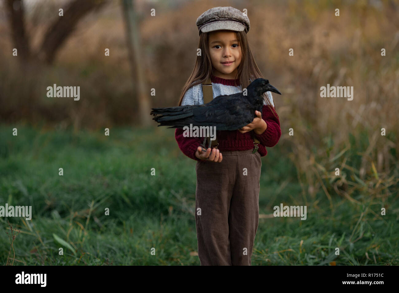 Child girl in cap stands with black raven in her hands on background of ...