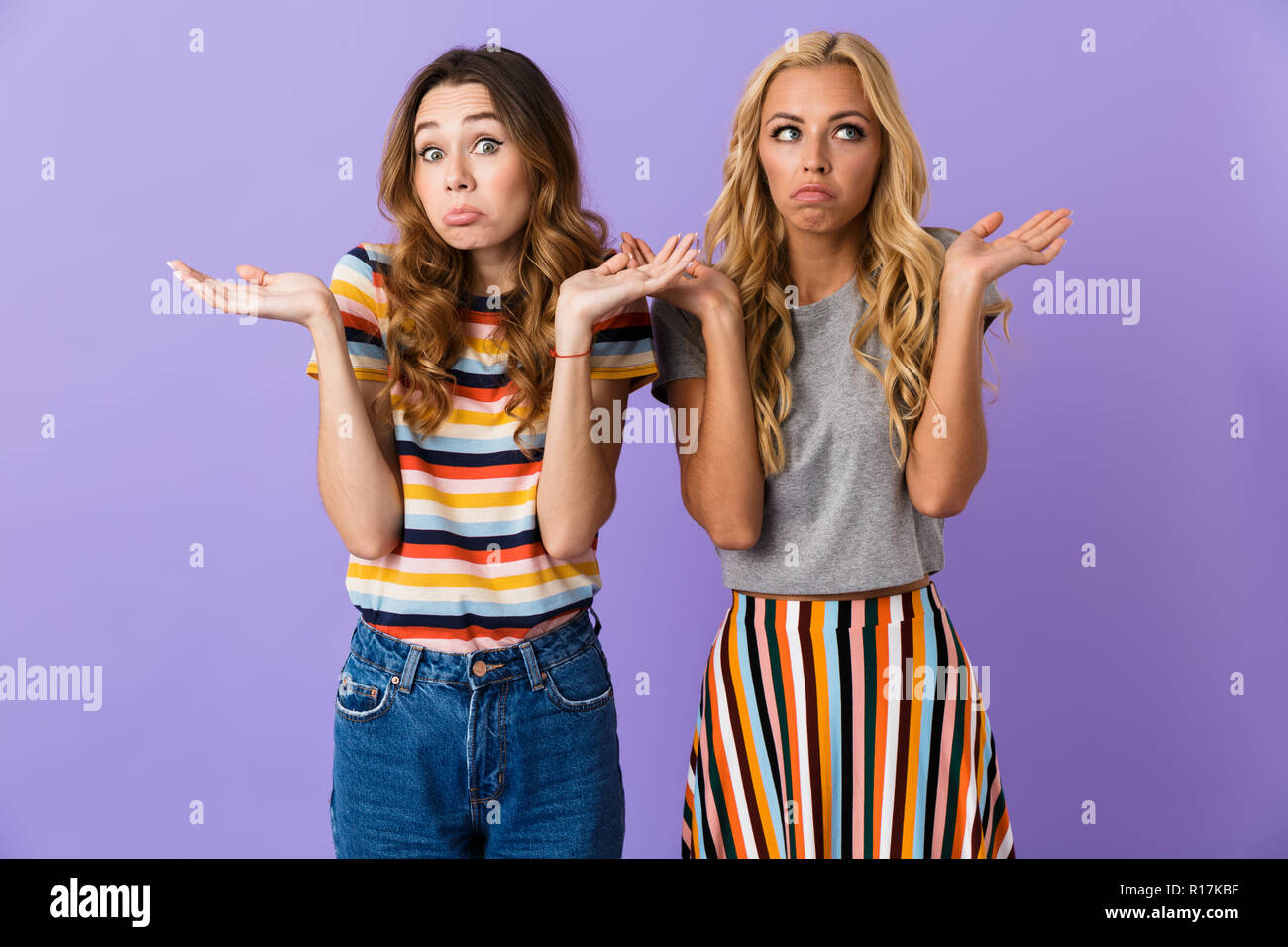 Two pretty confused young girls friends standing isolated over violet ...