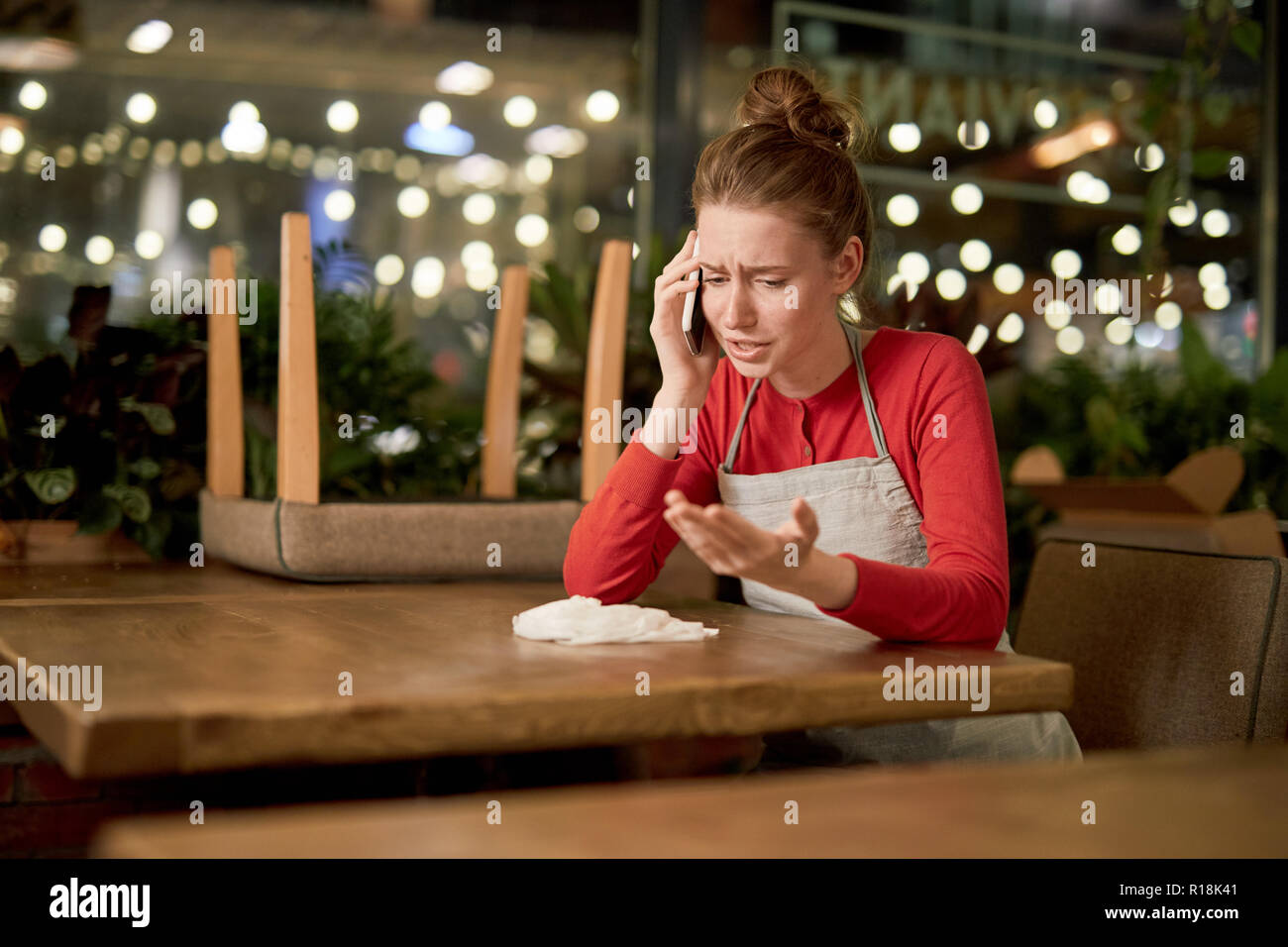 Upset waitress sitting by table and talking to someone by smartphone at ...