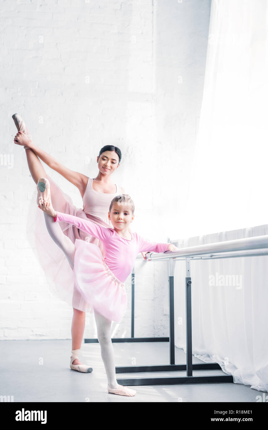 smiling young ballet teacher looking at little student stretching and ...