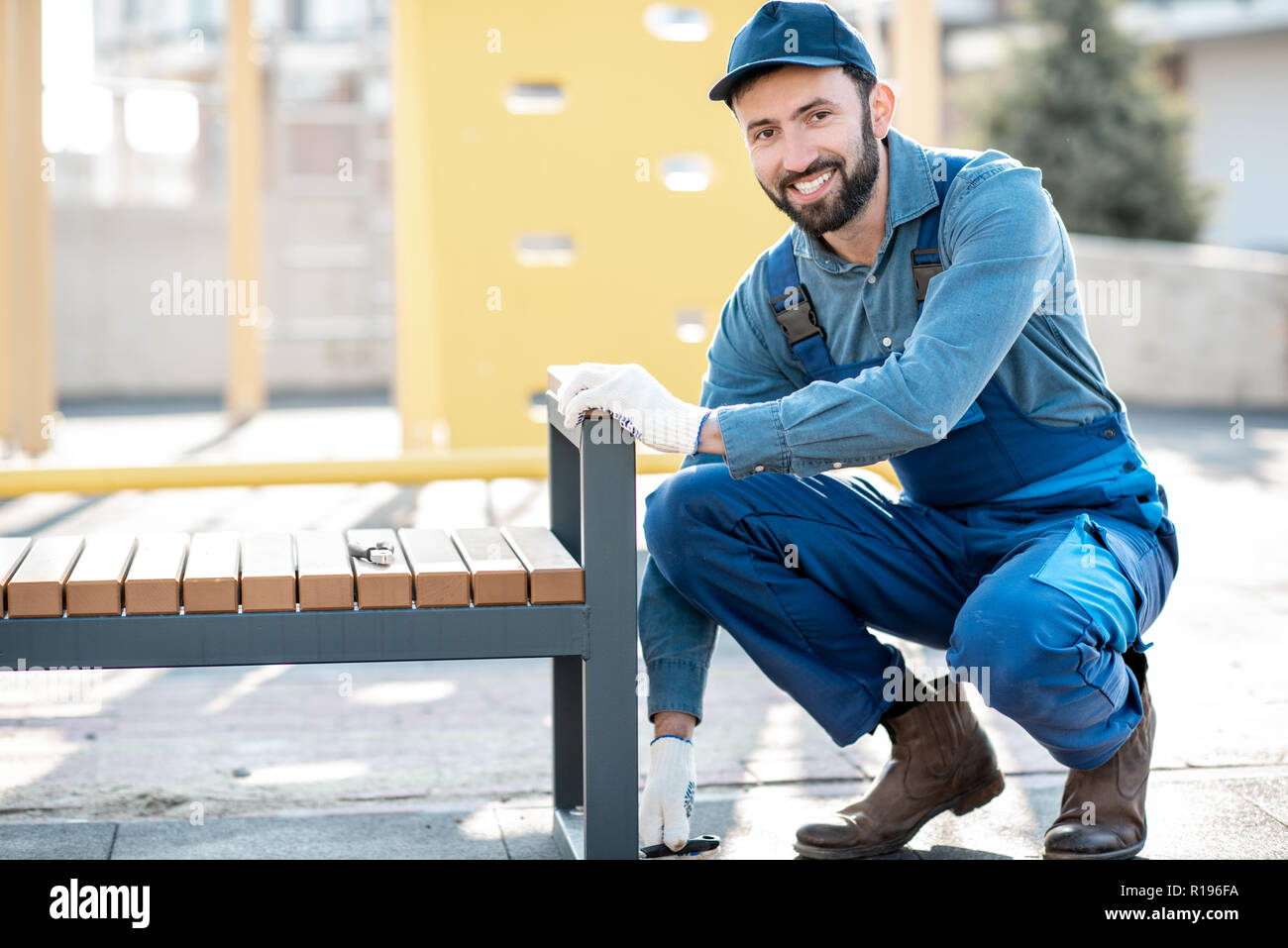 Handsome workman in uniform mounting a new bench on the playground ...