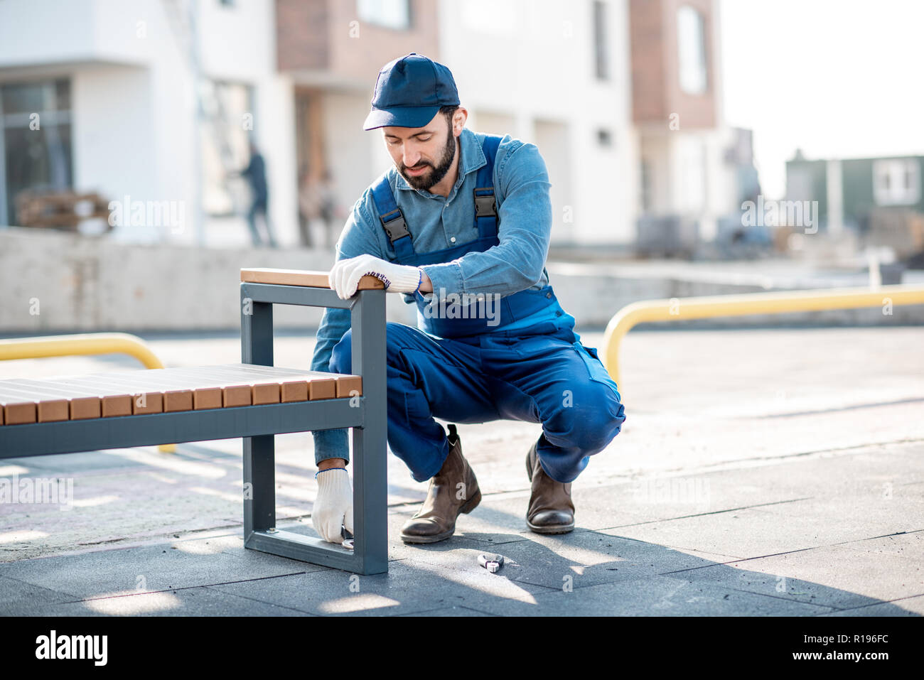 Handsome workman in uniform mounting a new bench on the playground ...