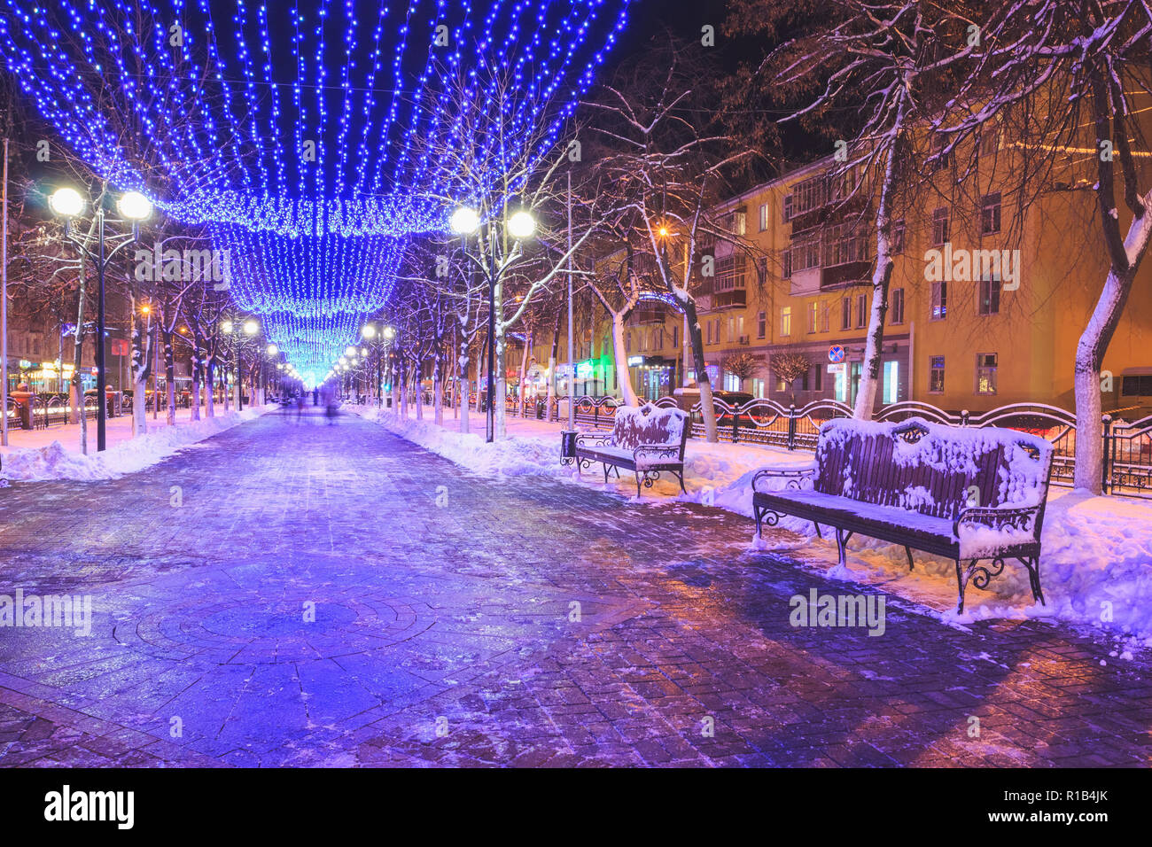 Alley in the city in the winter with benches in the snow and garlands ...
