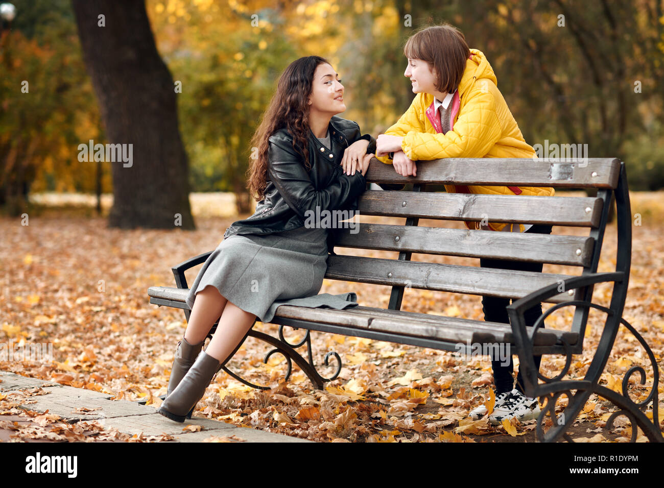 Two girls are in autumn city park. They sit on the bench and talk Stock ...