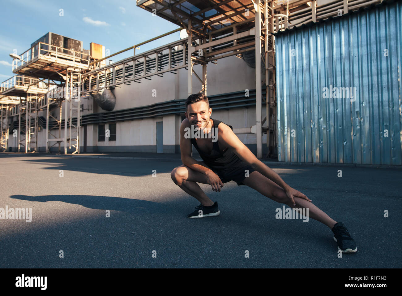 Smiling man doing warm up and stretching outdoor on in industrial city ...