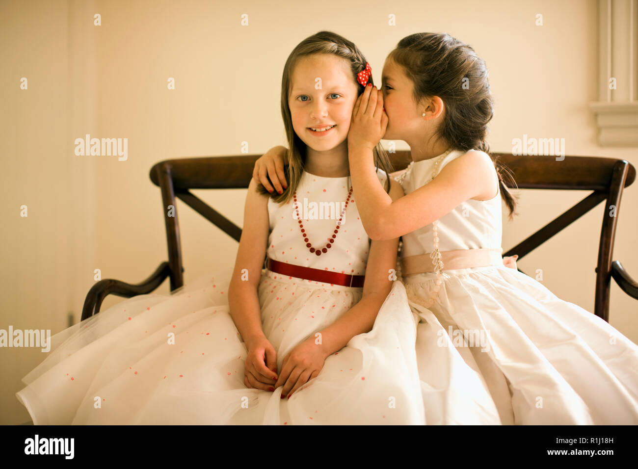 Young girls in white party dresses sit on wooden bench and whisper ...