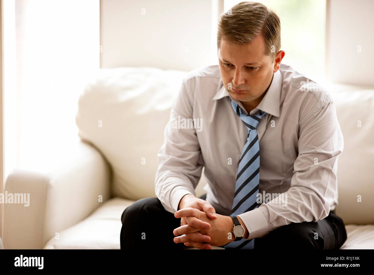 Despondent businessman sits on a couch with his hands clasped and looks ...