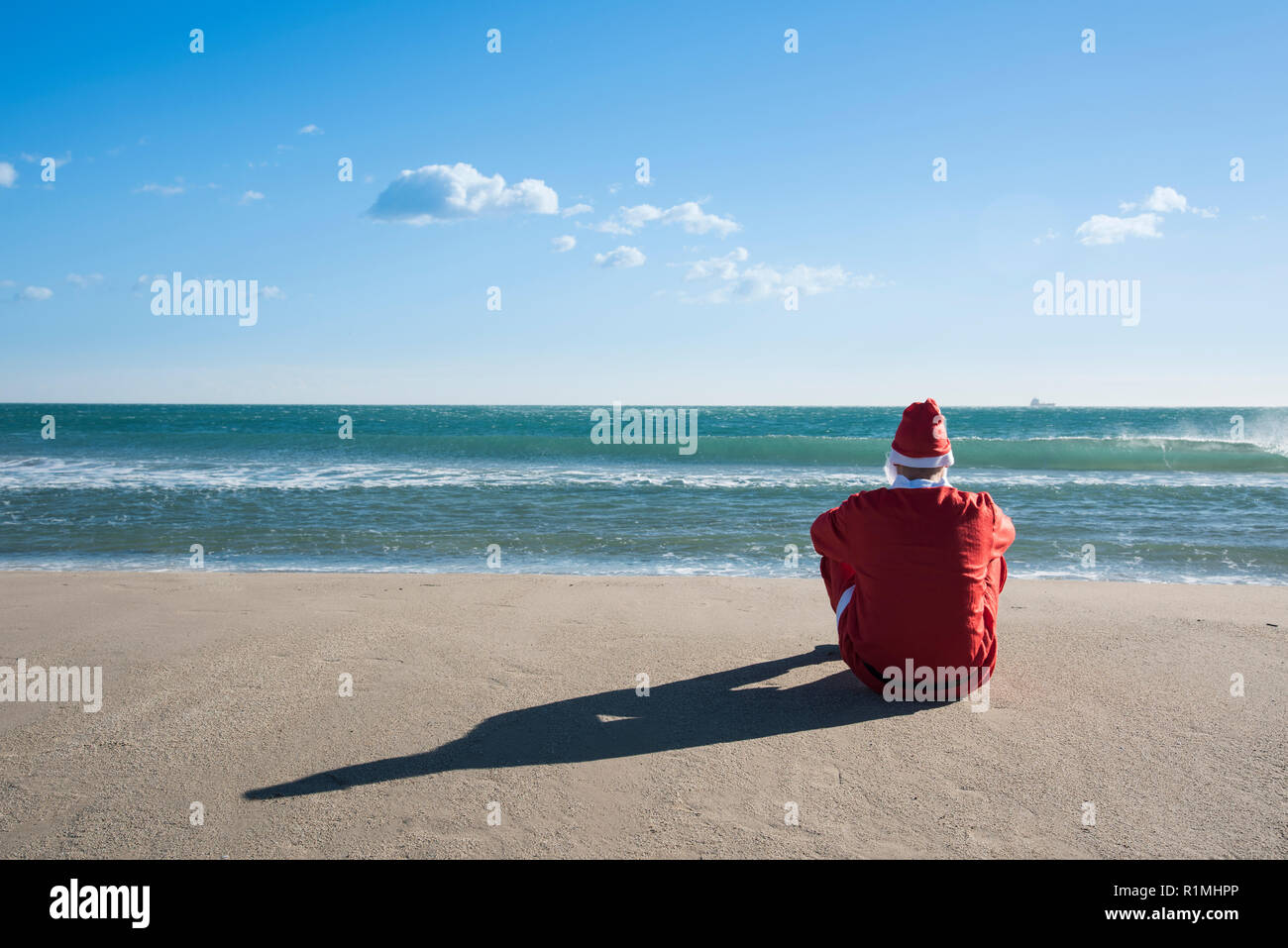 santa claus, seen from behind, sitting on the sand of a beach facing ...
