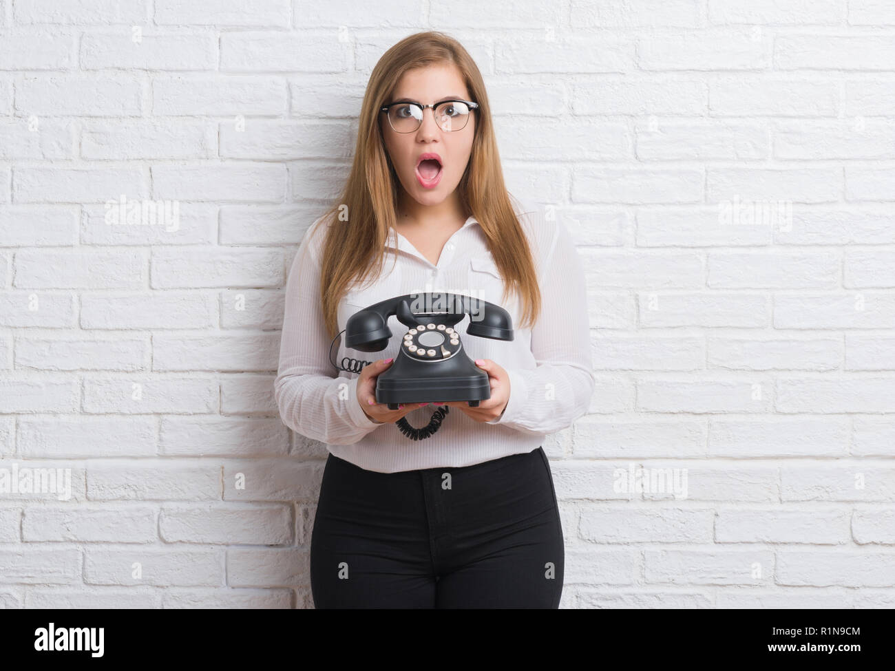 Young adult woman over white brick wall holding vintage telephone ...