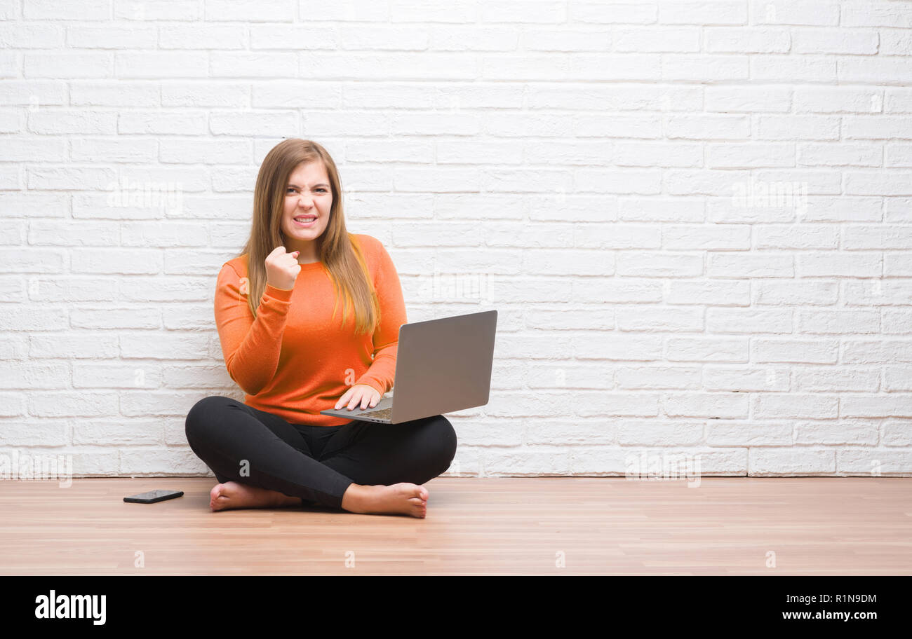 Young adult woman sitting on the floor over white brick wall using ...