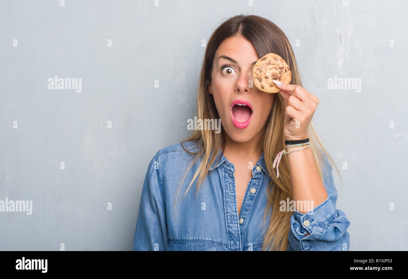 Young adult woman over grey grunge wall eating chocolate chip cooky ...