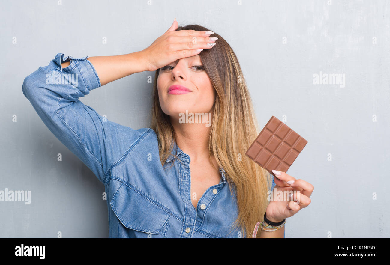 Young adult woman over grey grunge wall eating chocolate bar stressed ...