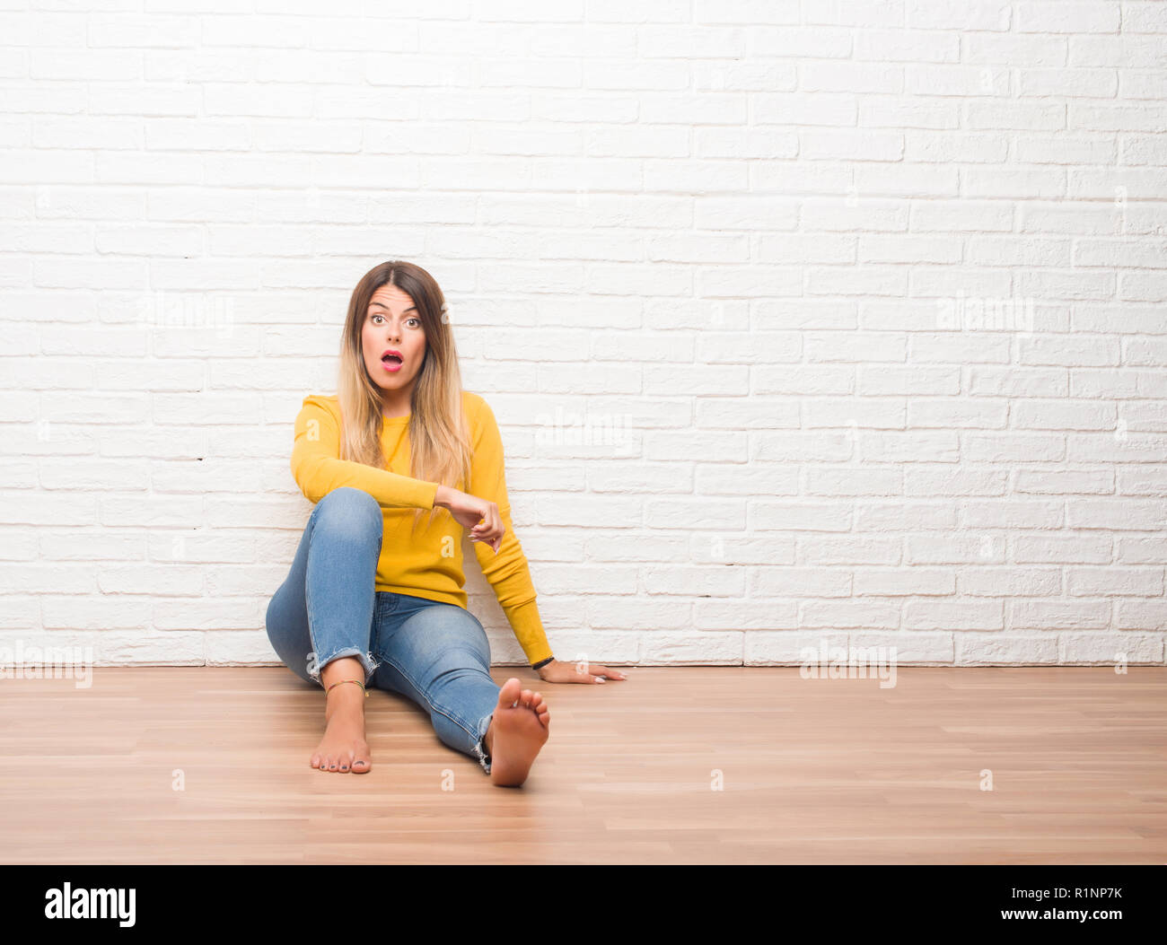 Young Adult Woman Sitting On The Floor Over White Brick Wall Afraid And