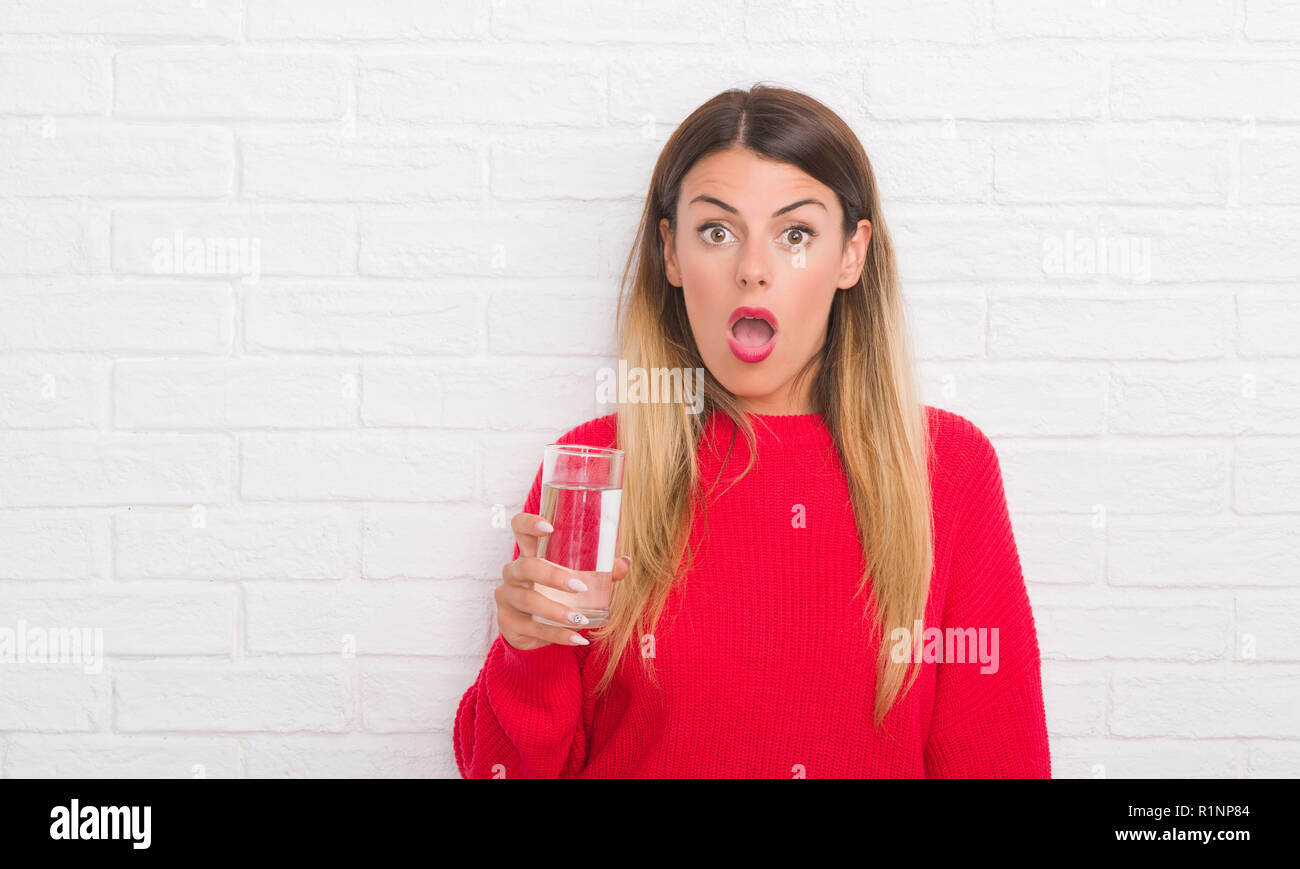 Young adult woman over white brick wall drinking glass of water scared ...