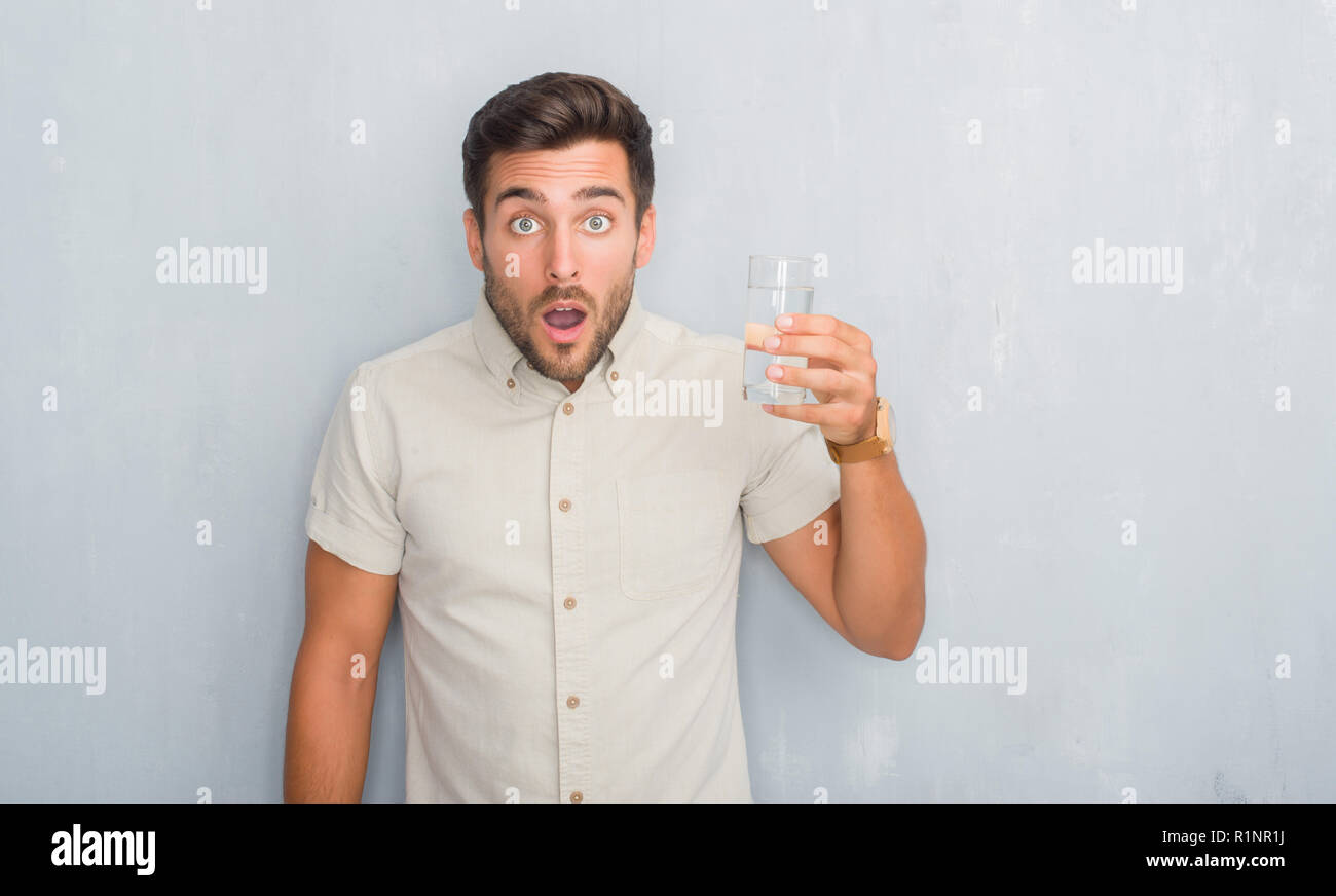 Handsome young man over grey grunge wall drinking glass of water scared ...