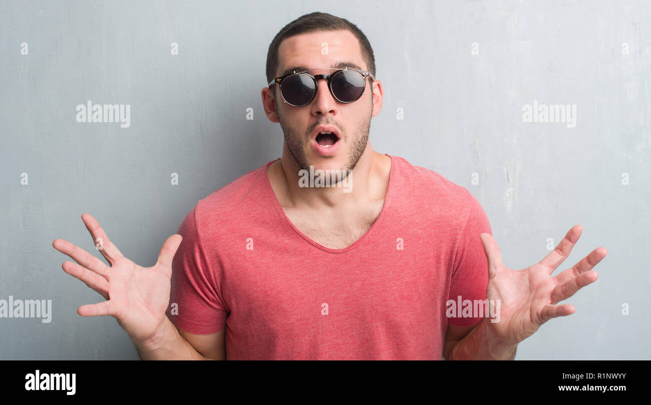 Young caucasian man over grey grunge wall wearing sunglasses scared in ...