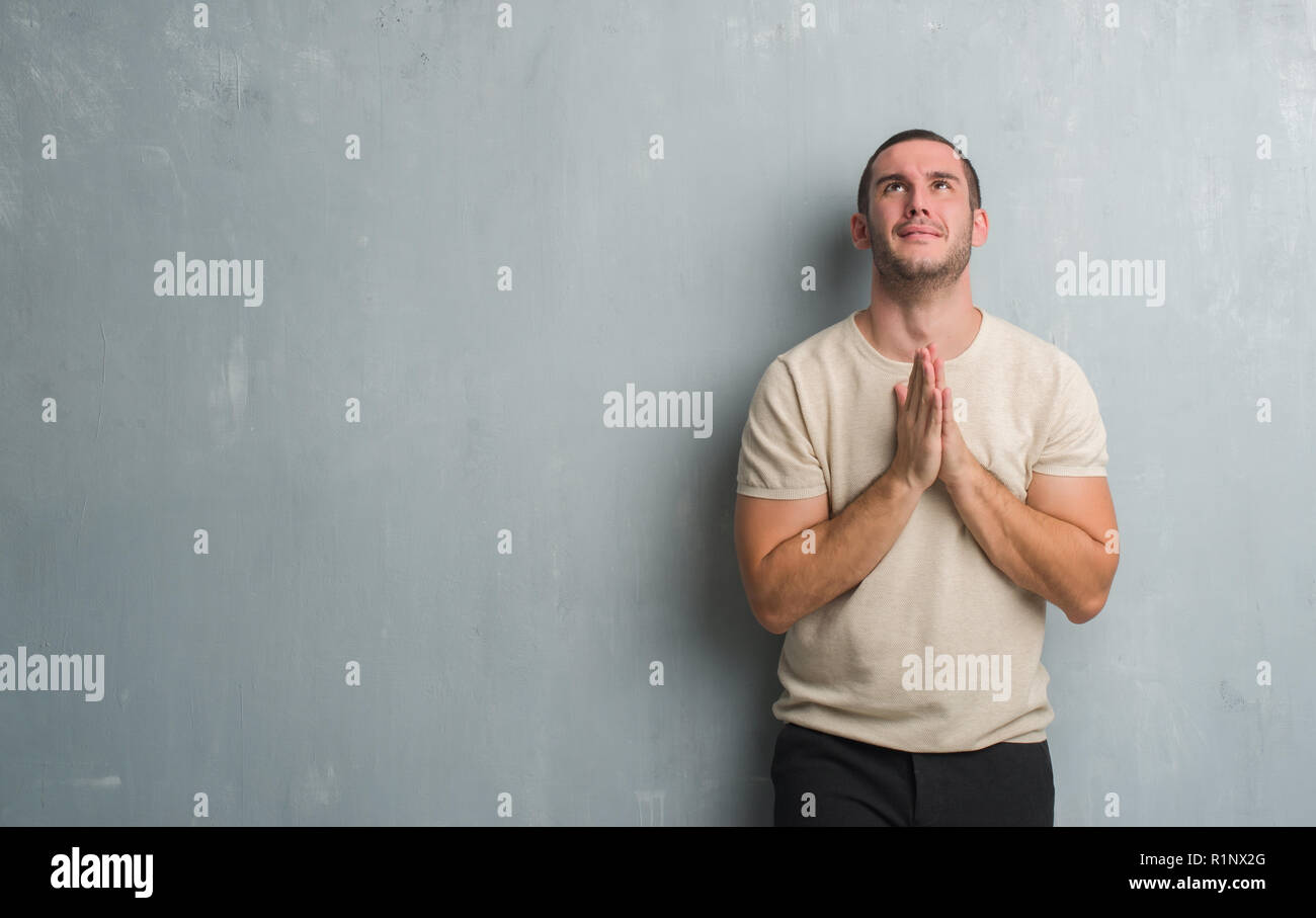 Young caucasian man over grey grunge wall begging and praying with ...