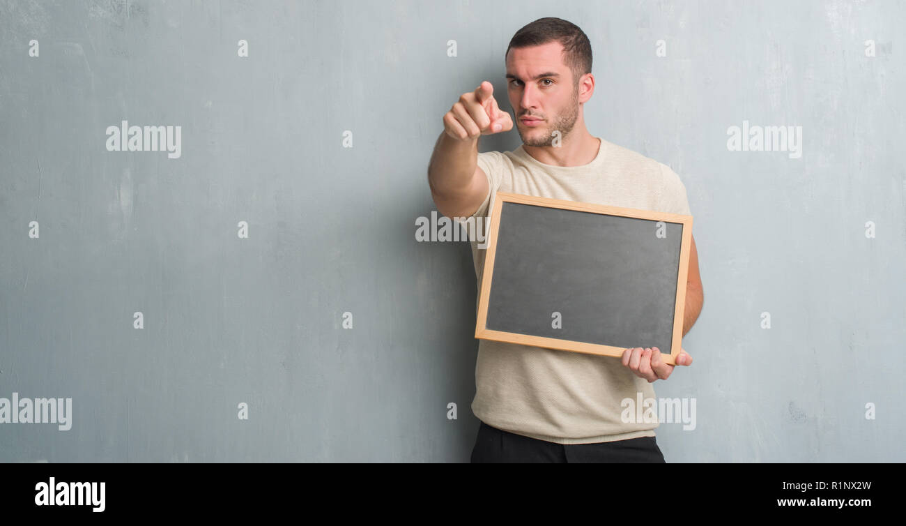 Young caucasian man over grey grunge wall holding blackboard pointing ...