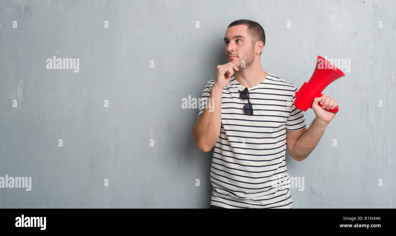 Young caucasian man over grey grunge wall shouting trough megaphone ...